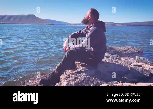 Young attractive traveler sitting on the rocky shore of a mountain lake ...