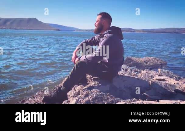 Young attractive traveler sitting on the rocky shore of a mountain lake ...