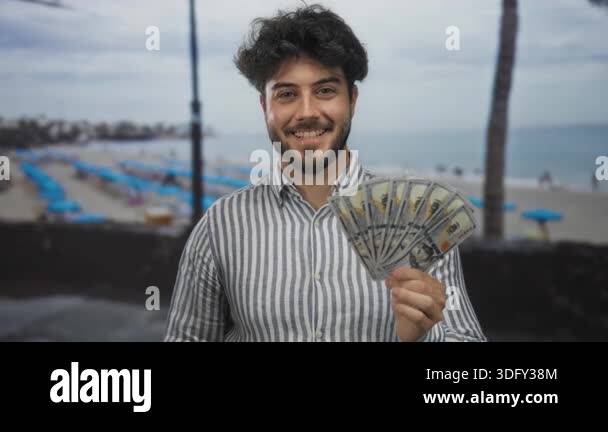 Young man with a beard holding us dollars, smiling at the seaside, with ...