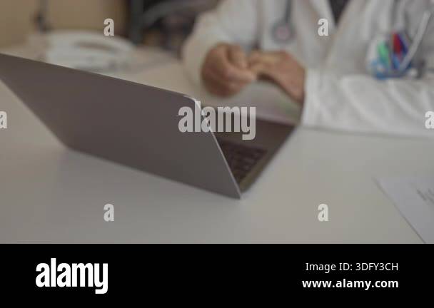 Doctor in white coat with stethoscope at desk in clinic using laptop ...