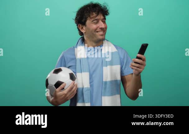 Hispanic man holding football and remote on green background, smiling ...