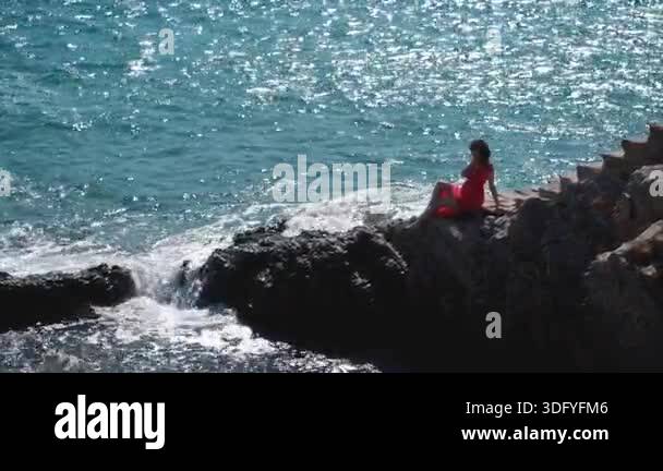 Girl in red dress sitting at ocean cliff beach Stock Video Footage - Alamy