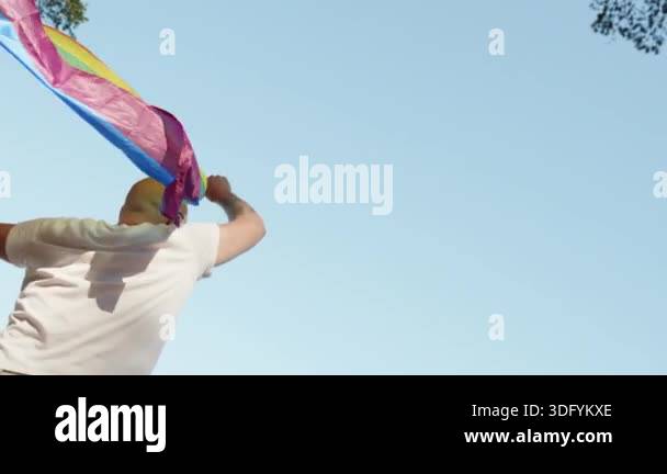 Queer man holding rainbow gay flag while parade on background of blue ...