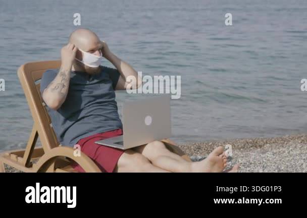 Man freelancer in medical mask work on laptop by the sea. Distant ...