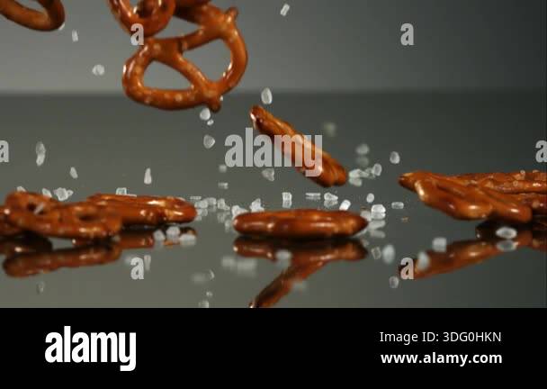 Several brown baked pretzel snacks fall onto a dark reflective surface ...
