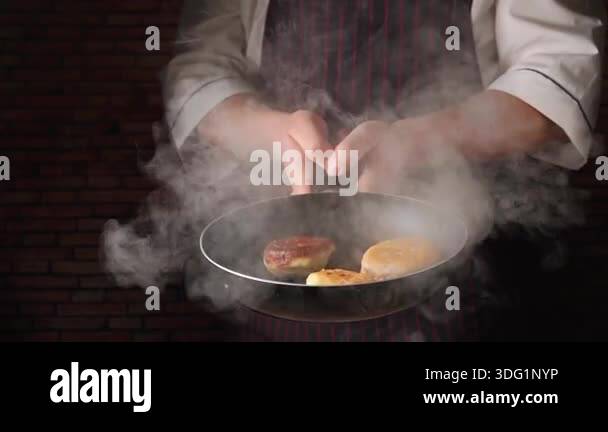 A chef stands in the kitchen, tossing a frying pan of doughnuts. Steam ...