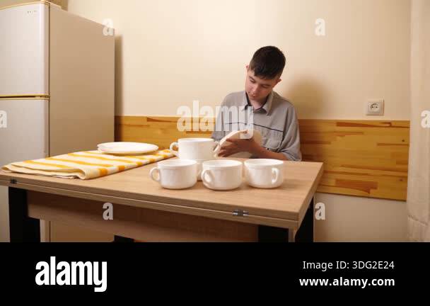 Teenager in a home kitchen drying and arranging clean dishes on a table ...