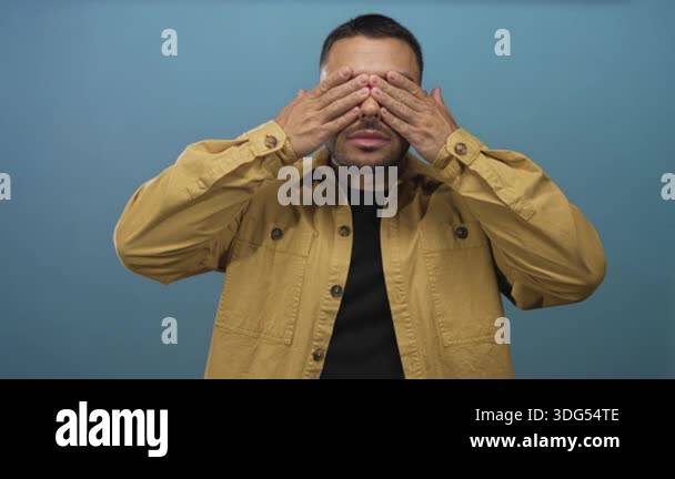 Young hispanic man in mustard jacket covers eyes with hands against ...