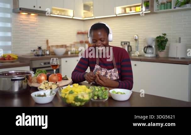 Happy woman wearing headphones is cutting fresh vegetables in a cozy ...