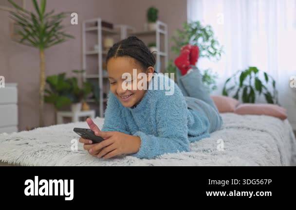 Young girl with braids lying on her bed, smiling while texting or ...
