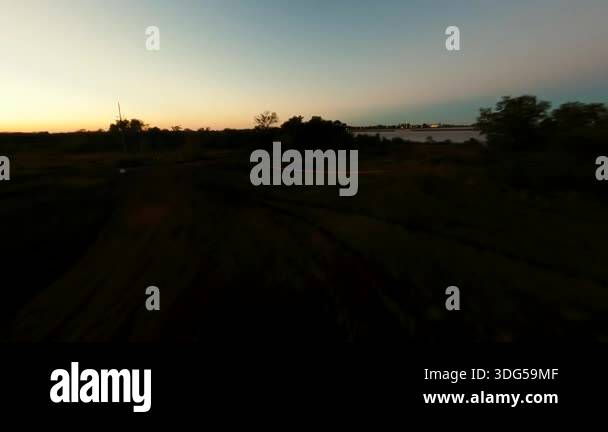 Fast FPV-style drone flight over a grassy riverside area at sunset ...