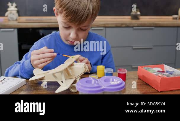 Focused boy applies paint with brush onto toy airplane wings sitting at ...