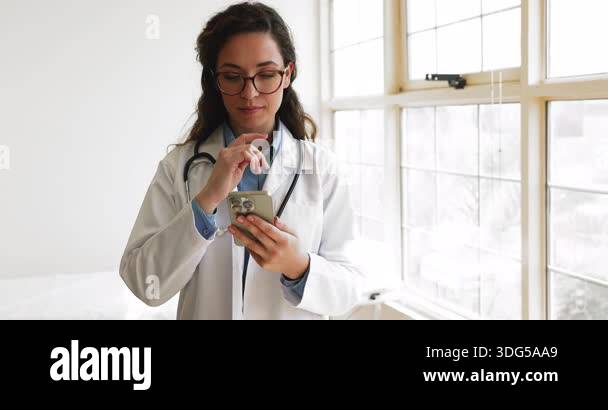 Smiling female doctor in white uniform using smartphone at workplace ...