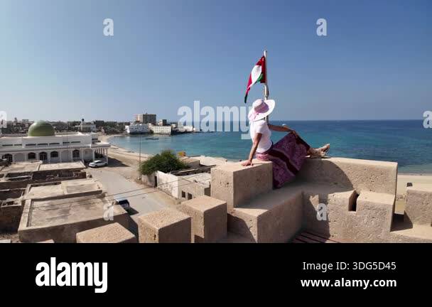 Woman in a hat and skirt sits on ancient Mirbat fort walls, overlooking ...