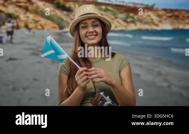 Smiling woman holding scottish flag on seaside beach, wearing a hat ...