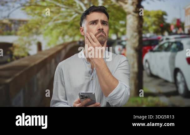 Young hispanic man with smartphone pondering on a city street lined ...