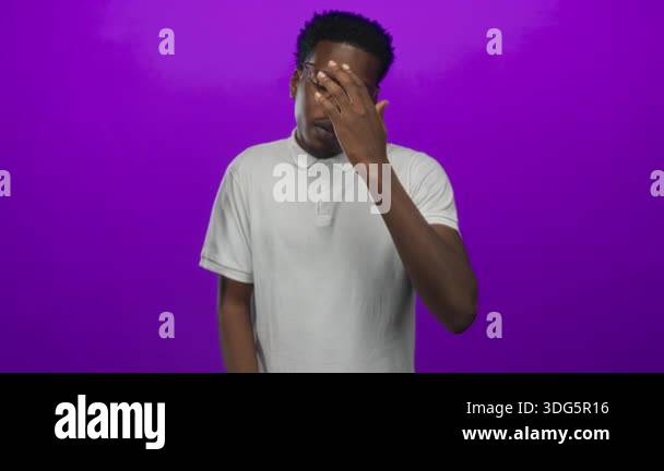 Young man with glasses covers his face with one hand in purple studio ...