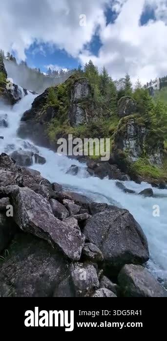A vibrant white waterfall plunges from a high rocky cliff into a fast ...