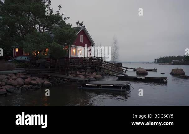 Red wooden villa with warm glowing windows stands on rocky shore at ...
