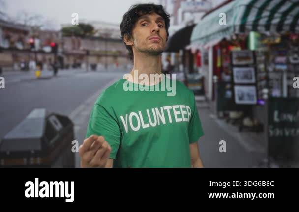 Young man wearing green volunteer shirt beckons with open hand on city ...