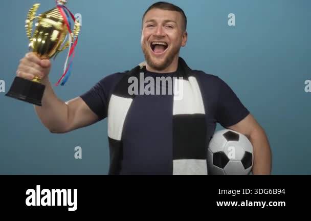Man holding trophy and soccer ball shouting in blue studio wearing ...