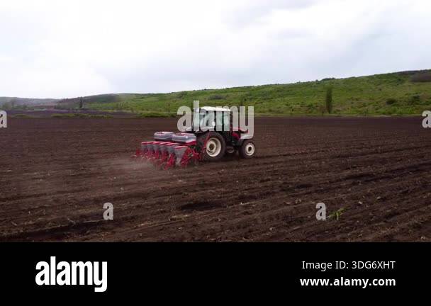 A red tractor equipped with a seed planter advances through a plowed ...