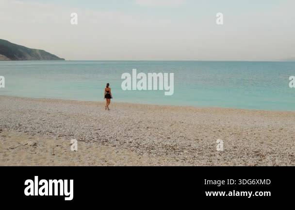 Woman walking beside calm turquoise sea along wide pebble beach on a ...