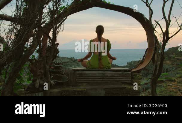 Woman in green dress meditates in wooden structure with ocean sunset ...