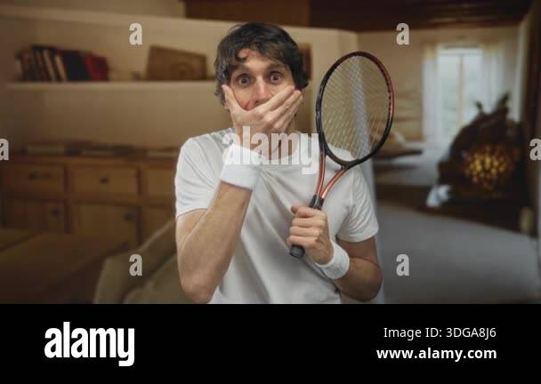 Wide eyed young man in white shirt and wristbands holds tennis racket ...