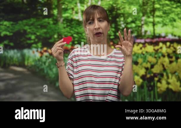 Woman holds small red heart with left hand in forest, lips pursed in ...