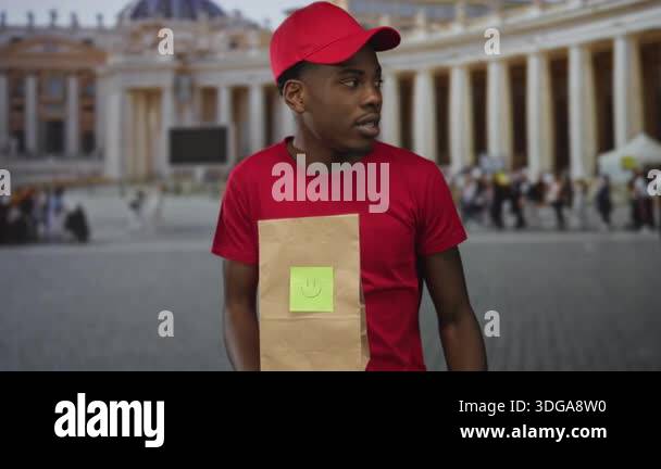 Man in red cap and shirt holds paper bag while shading eyes with raised ...