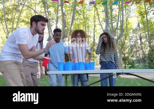 Multiethnic friends enjoying competitive beer pong game in sunlit park ...