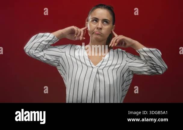 Woman plugging ears with fingers in studio against a deep red backdrop ...
