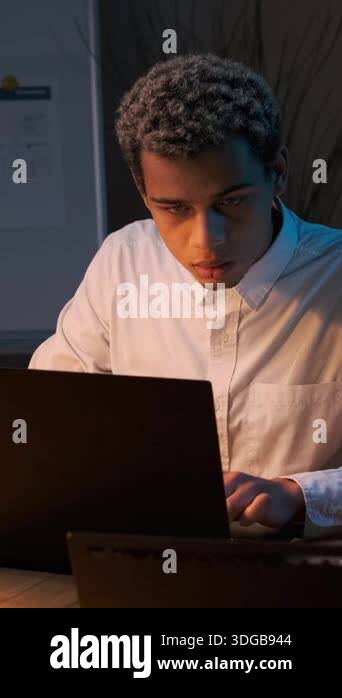 Vertical video of a tired young man working late on his laptop in a ...