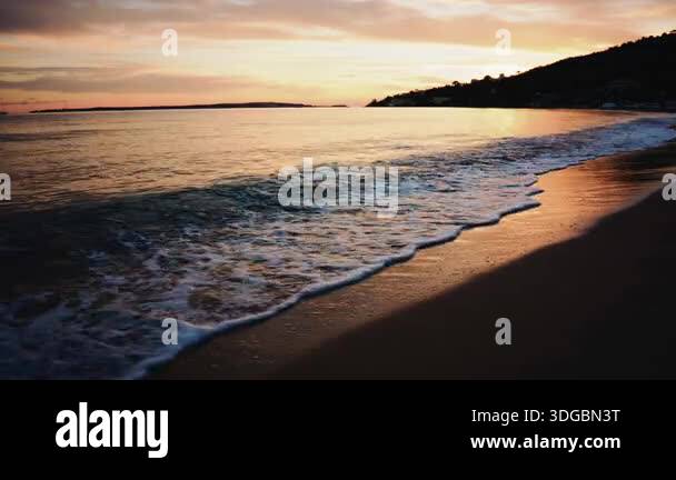 Gentle sea waves rolling onto a sandy beach during sunset, with ...