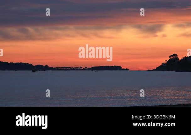 Wide shot of calm sea under a colorful sunset sky, with distant islands ...