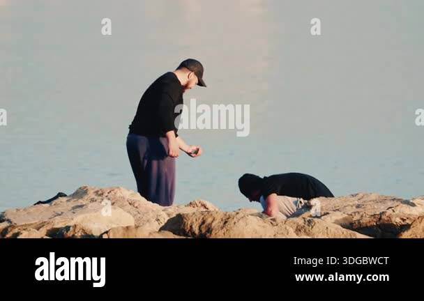 Antibes, France - December 22, 2025: Two men crouching on coastal rocks ...