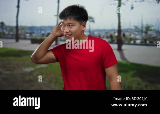 Young chinese man in red shirt smiles and listens outdoors on a city ...