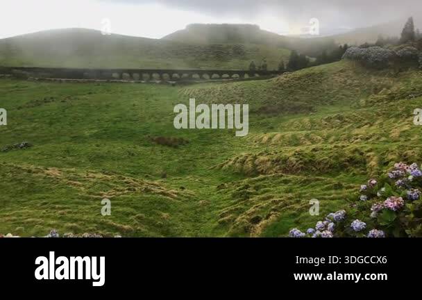 A person stands in a vast green field surrounded by hills. A stone ...