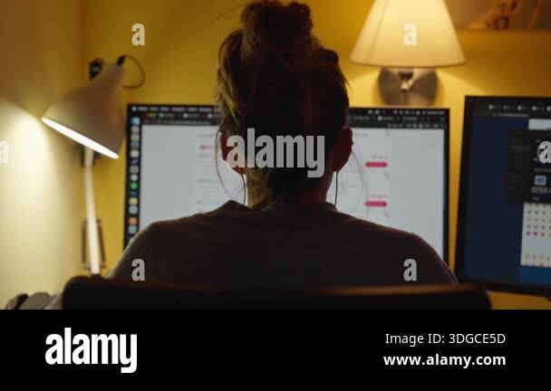 Young woman from behind using a computer with two monitors in a dimly ...
