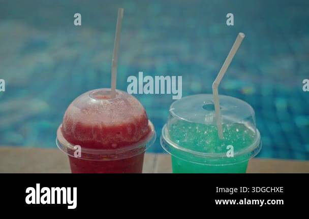 Two frozen tropical drinks in plastic cups with straws on pool edge ...