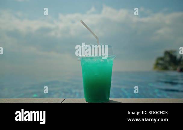 Green frozen tropical drink in plastic cup with straw on pool edge ...