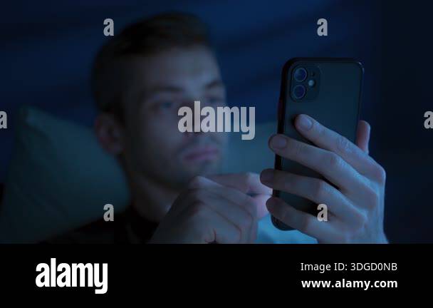 A young man lies alone in a dark bedroom, holding mobile phone and ...