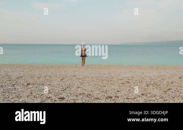 Woman facing the horizon while walking on a quiet pebble beach with ...