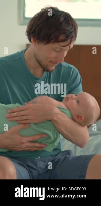 A father gently rocks his swaddled newborn baby to sleep while sitting ...