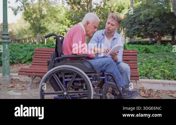 Young man sitting on a bench in a park, reading a book to his elderly ...