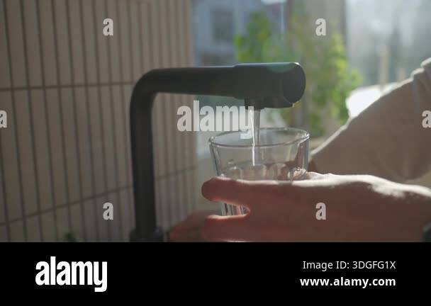 Hand holding glass under black kitchen faucet with sunlight from window ...