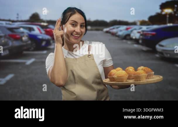 Young woman baker in apron holding a wooden tray of muffins and points ...
