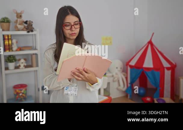 Young hispanic woman teacher reading a book inside a kindergarten ...