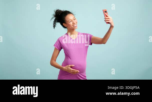 Smiling pregnant woman taking a selfie and caressing her belly against ...